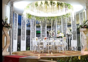 Uma área de estar circular com cadeiras e mesas brancas, rodeada por plantas pendentes decorativas e arranjos florais.