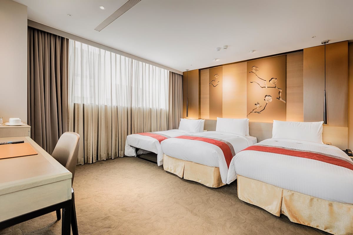 Quarto de hotel com duas camas, uma mesa de trabalho e uma janela grande com cortinas.