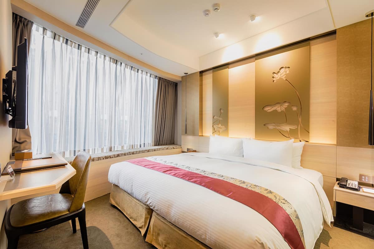 Quarto de hotel com uma cama grande, uma escrivaninha com cadeira, uma televisão e uma janela com cortinas.