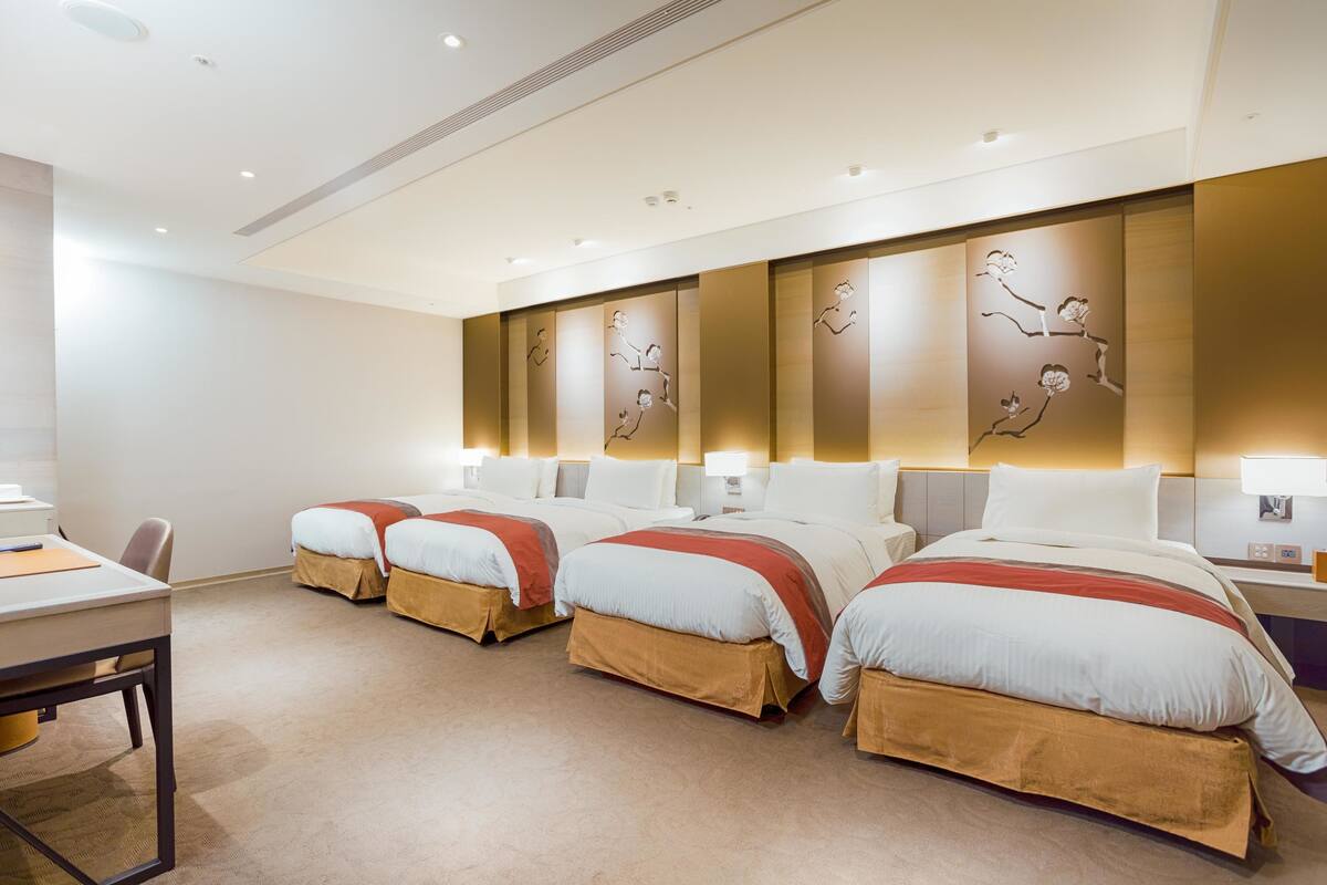 Quarto de hotel com três camas, uma escrivaninha e uma parede grande com painéis decorativos.