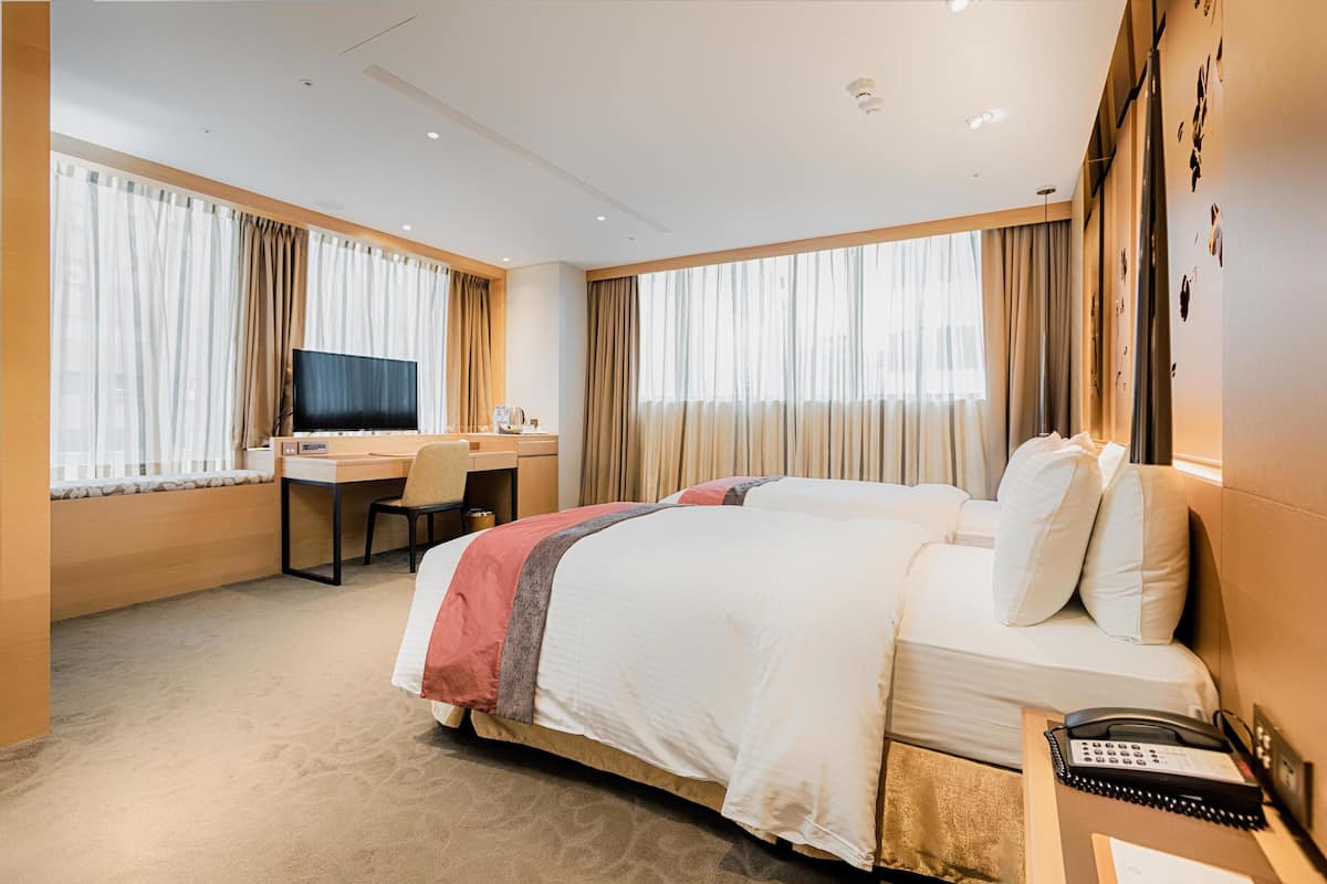 Quarto de hotel com duas camas, uma escrivaninha com cadeira, televisão e telefone em uma mesinha de cabeceira.