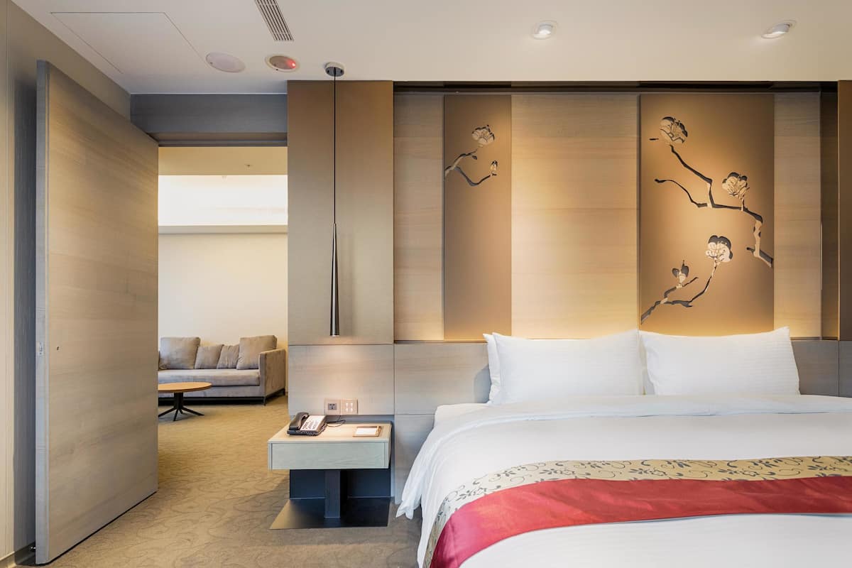 Quarto de hotel moderno com uma cama grande, mesa de cabeceira com telefone, sofá e um painel decorativo na parede.
