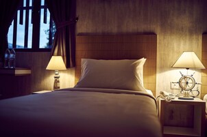 Quarto de hotel com uma cama grande, duas luminárias de cabeceira, mesa de cabeceira com telefone e um objeto decorativo, além de uma janela com cortinas.