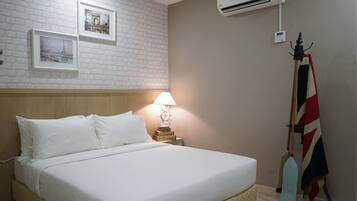 Cama bem arrumada com lençóis brancos, cabeceira de madeira e ar-condicionado fixado na parede. Há duas molduras com quadros na parede, e um remo de madeira apoiado na parede.