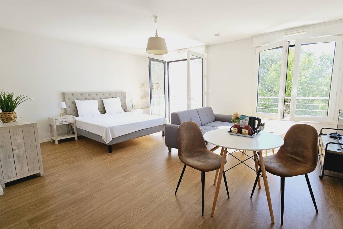 Quarto moderno e bem iluminado, com cama, sofá, duas cadeiras, mesa de jantar e pequena cozinha.
