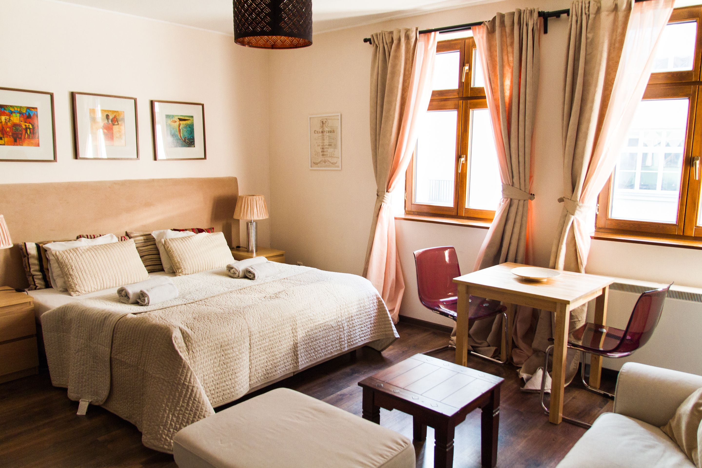 Quarto de hotel com uma cama grande, uma mesa de escritório, uma cadeira e um sofá. Há cortinas nas janelas e obras de arte nas paredes.