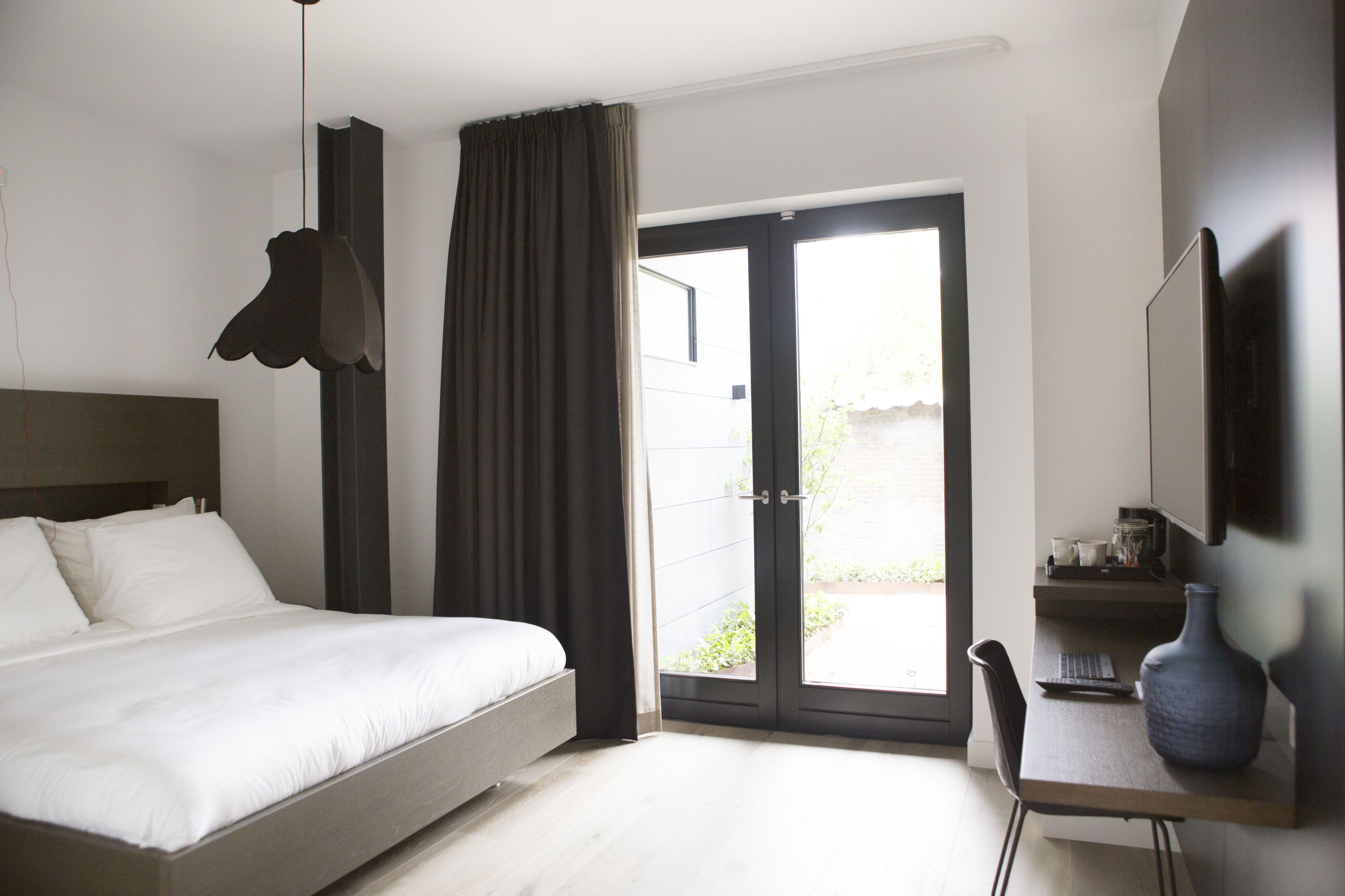 Quarto de hotel moderno com uma cama, escrivaninha, cadeira, janela grande e porta que dá para uma área externa.