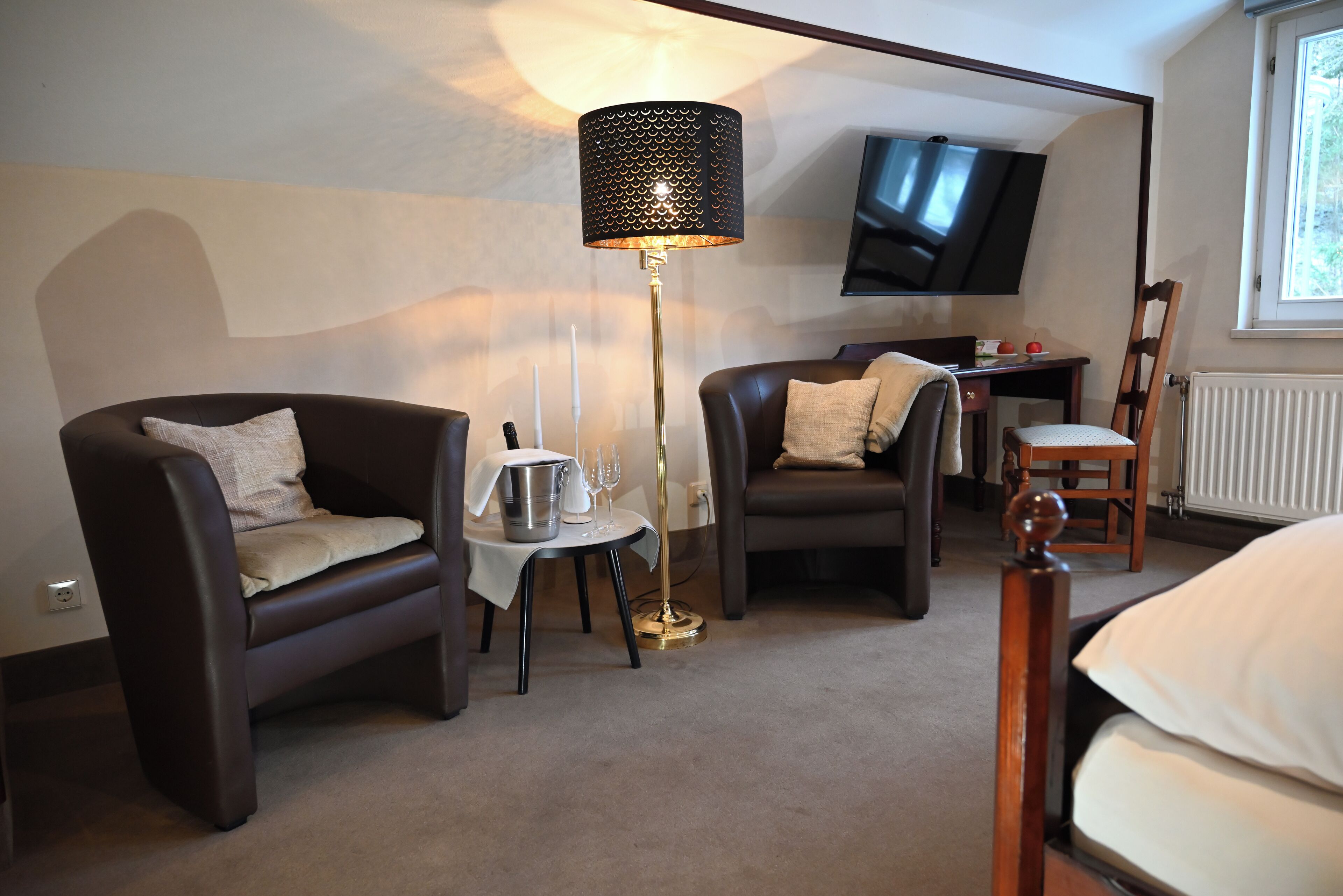 Quarto de hotel com sofá, poltrona, mesa de cabeceira, luminária, televisão e vista para o exterior.
