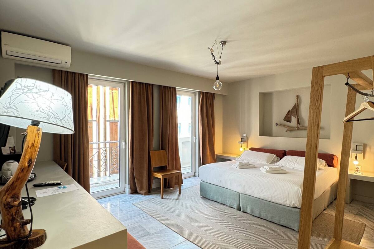 Quarto de hotel com cama, escrivaninha com luminária, cadeira, guarda-roupa e vista para o exterior.