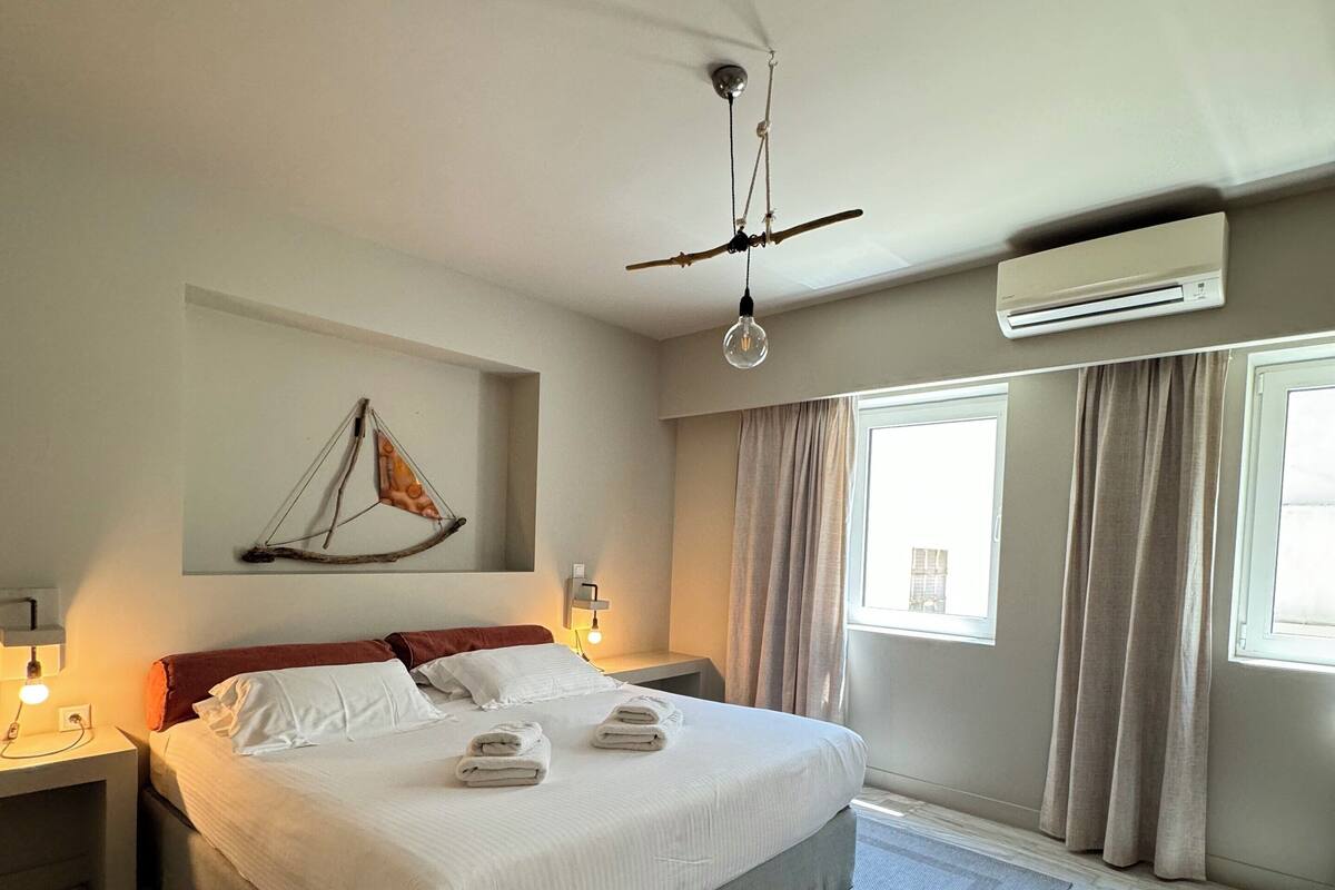 Quarto de hotel com uma cama grande, mesas de cabeceira com luminárias, um ventilador de teto, um aparelho de ar condicionado e uma janela com cortinas.