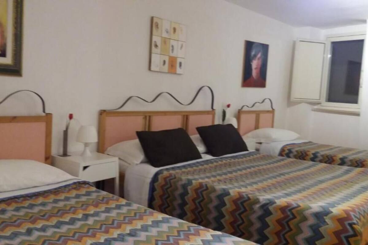 Quarto de hotel com três camas, cada uma com um distinto cobertor estampado em zigue-zague. Há mesinhas de cabeceira brancas com luminárias e uma prateleira na parede acima das camas.