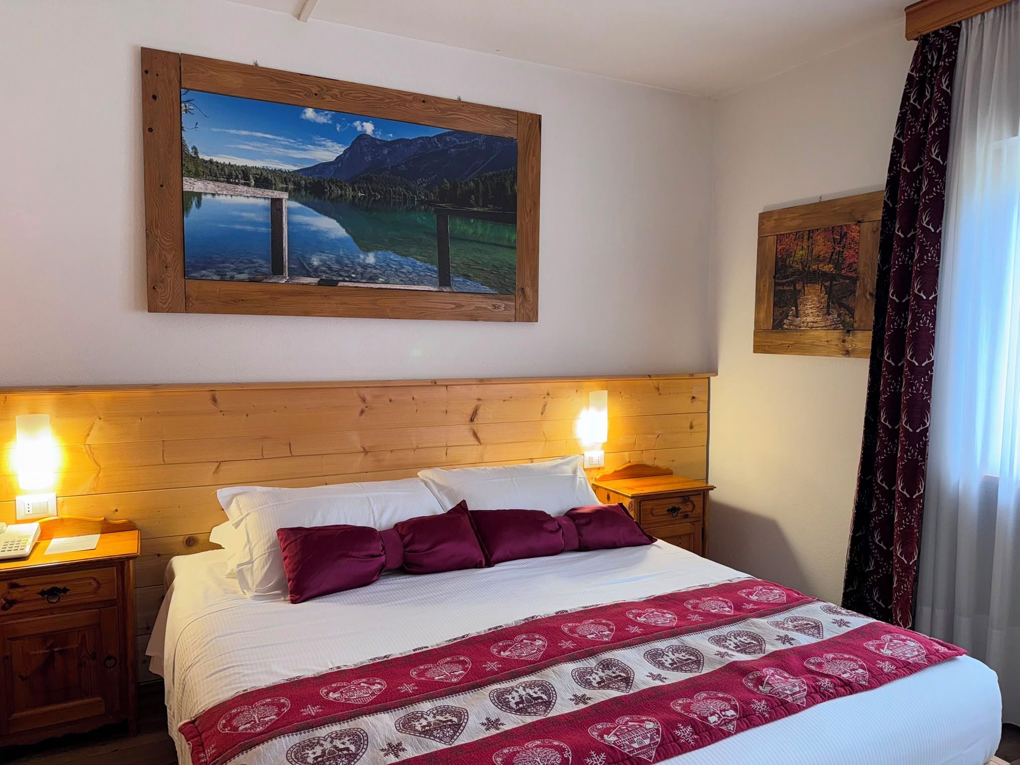 Quarto de hotel com cabeceira de madeira, uma cama grande com roupa de cama branca e um cobertor estampado em vermelho, duas luminárias de cabeceira e um quadro de paisagem acima da cama.