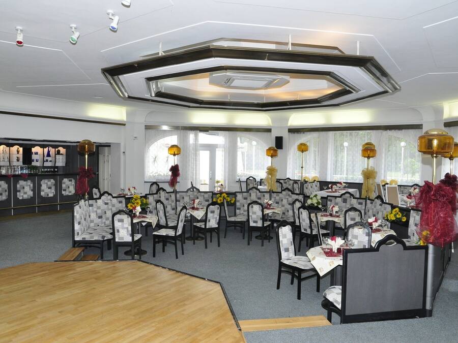 Uma sala de banquetes com pista de dança central, diversas mesas de jantar com cadeiras e um espaço bar com revestimento de azulejos.