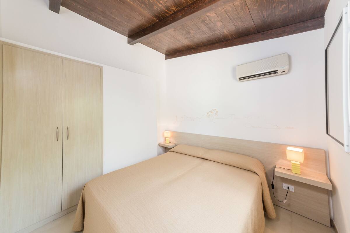 Quarto com cama, mesa de cabeceira, criado-mudo e unidade de ar condicionado na parede.