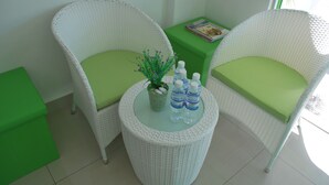 Quarto com duas cadeiras de vime brancas e almofadas verdes, uma mesa redonda pequena com uma almofada verde, uma planta em vaso e três garrafas de água.