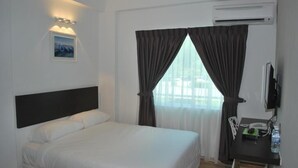 Quarto de hotel com cama, uma secretária com telefone e uma garrafa, e janela com cortinas.