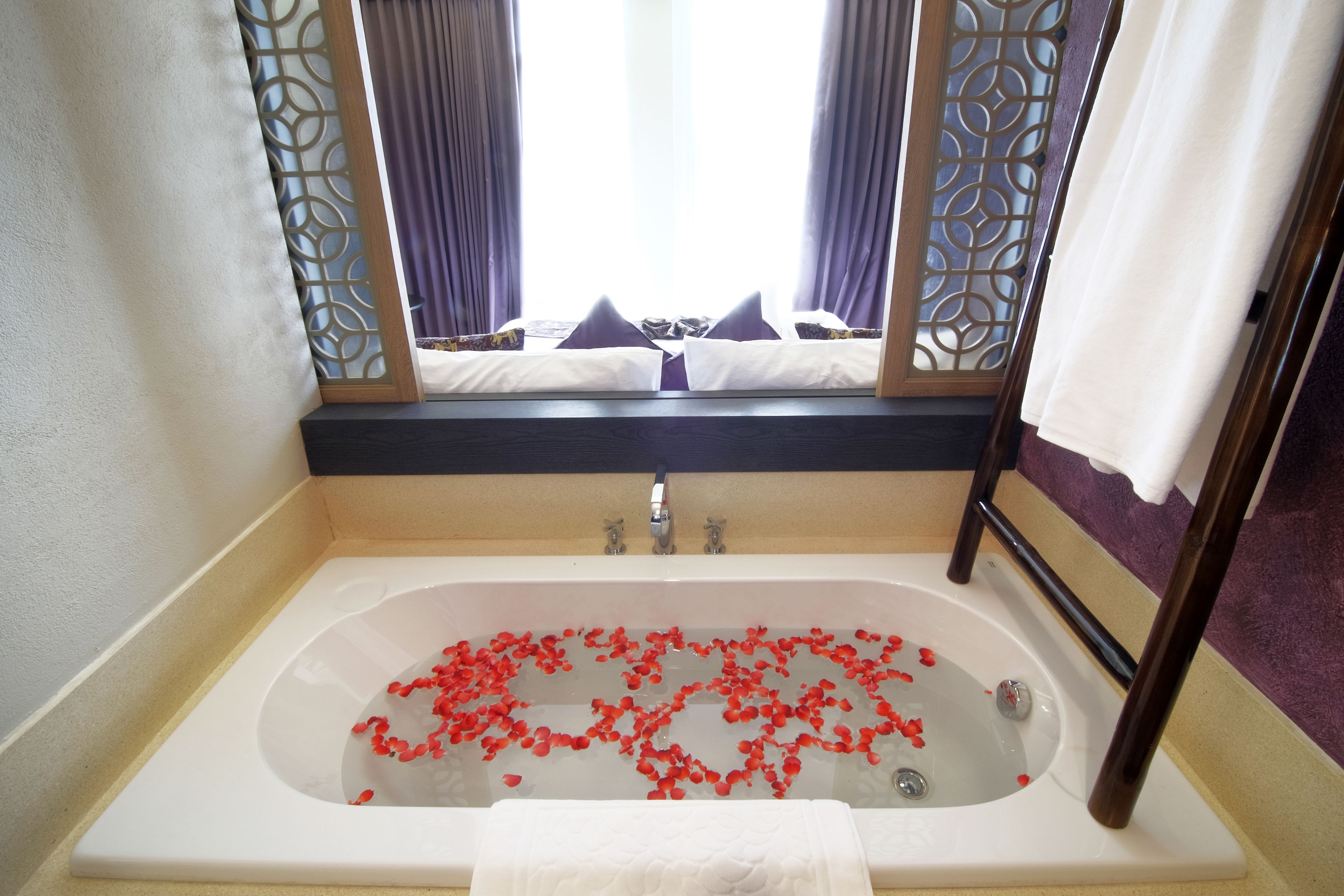 Um banheiro com banheira cheia de pétalas de rosa, janela com cortinas roxas e cama com roupa de cama branca.