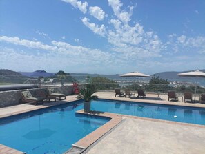 Área de piscina com espreguiçadeiras e guarda-sóis, com vista para uma paisagem deslumbrante com montanhas e um corpo d'água.