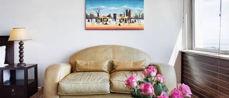 Uma sala de estar com um sofá bege, uma mesa pequena com abajur, um vaso de flores cor-de-rosa e um quadro na parede.