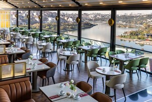 Restaurante com mesas e cadeiras dispostas para refeições, grandes janelas com vista para o rio e a paisagem urbana.
