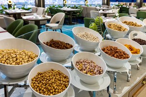 Buffet com diversos alimentos dispostos em recipientes sobre uma mesa, numa área de refeições bem iluminada com cadeiras e mesas verdes.