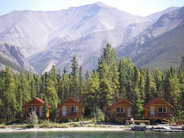 Uma fileira de cabanas de madeira com telhados vermelhos, localizadas à beira de um lago, com montanhas ao fundo.