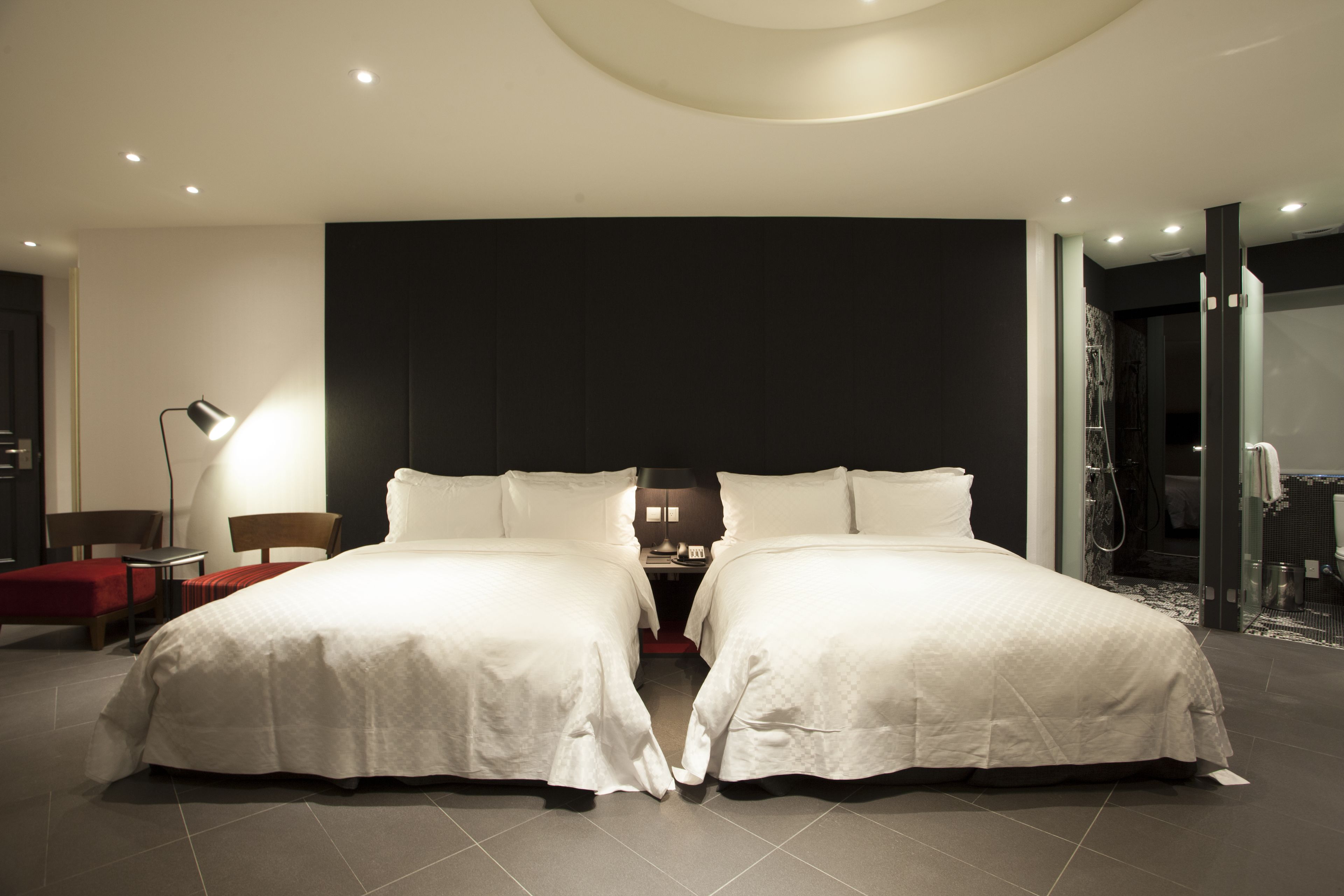 Quarto de hotel moderno com duas camas, uma escrivaninha, uma cadeira e um banheiro visível por uma porta aberta.