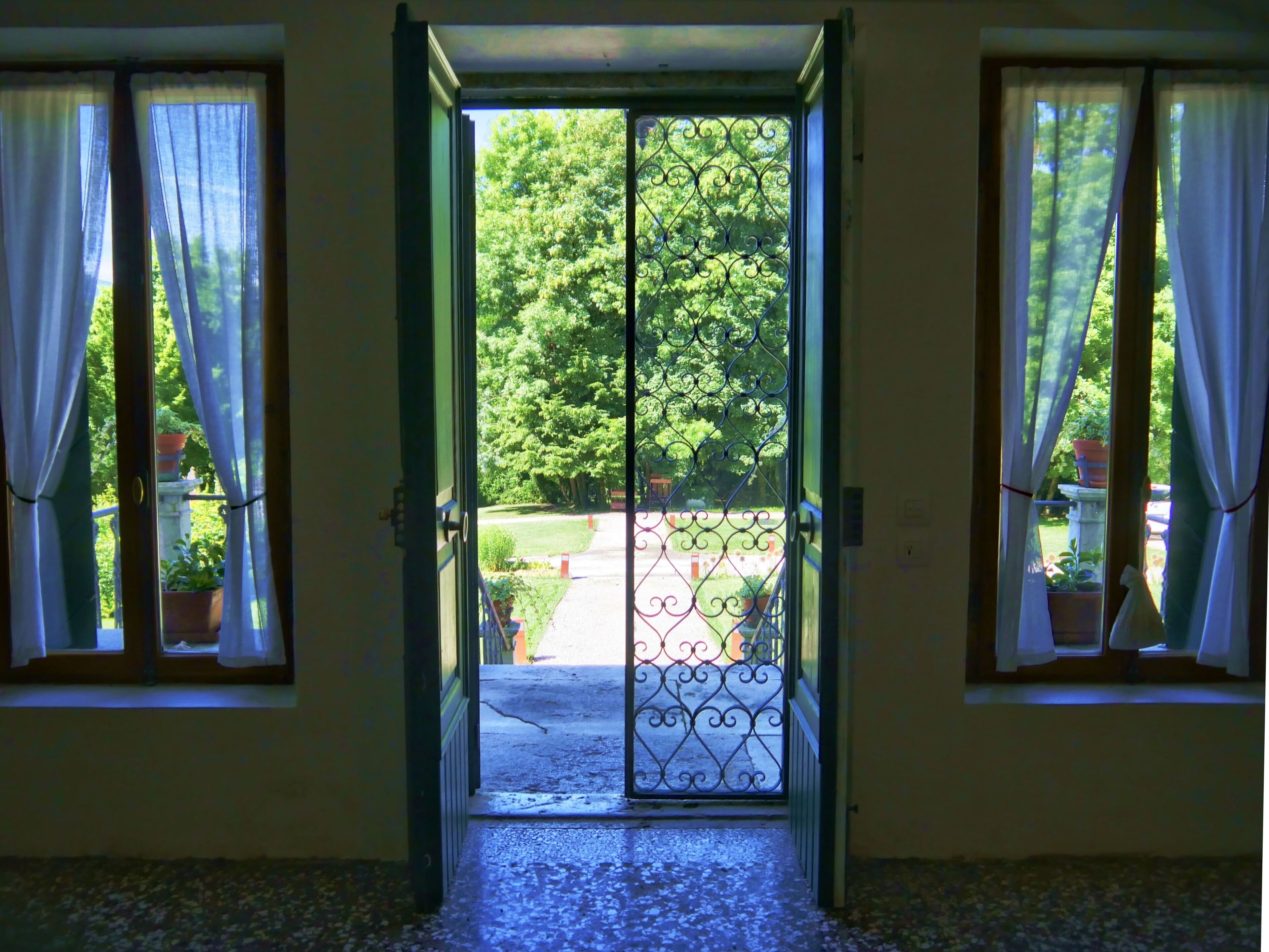Uma porta que dá para uma varanda com grade de metal, ladeada por janelas com cortinas.