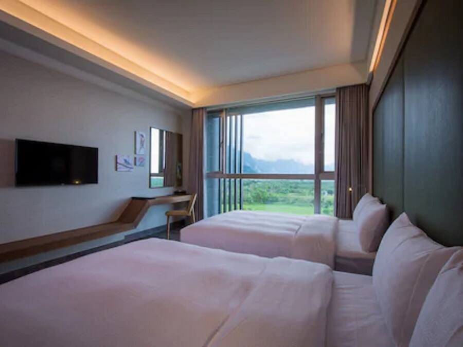 Quarto de hotel com duas camas, uma escrivaninha, TV de tela plana e uma grande janela com vista para vegetação e montanhas.