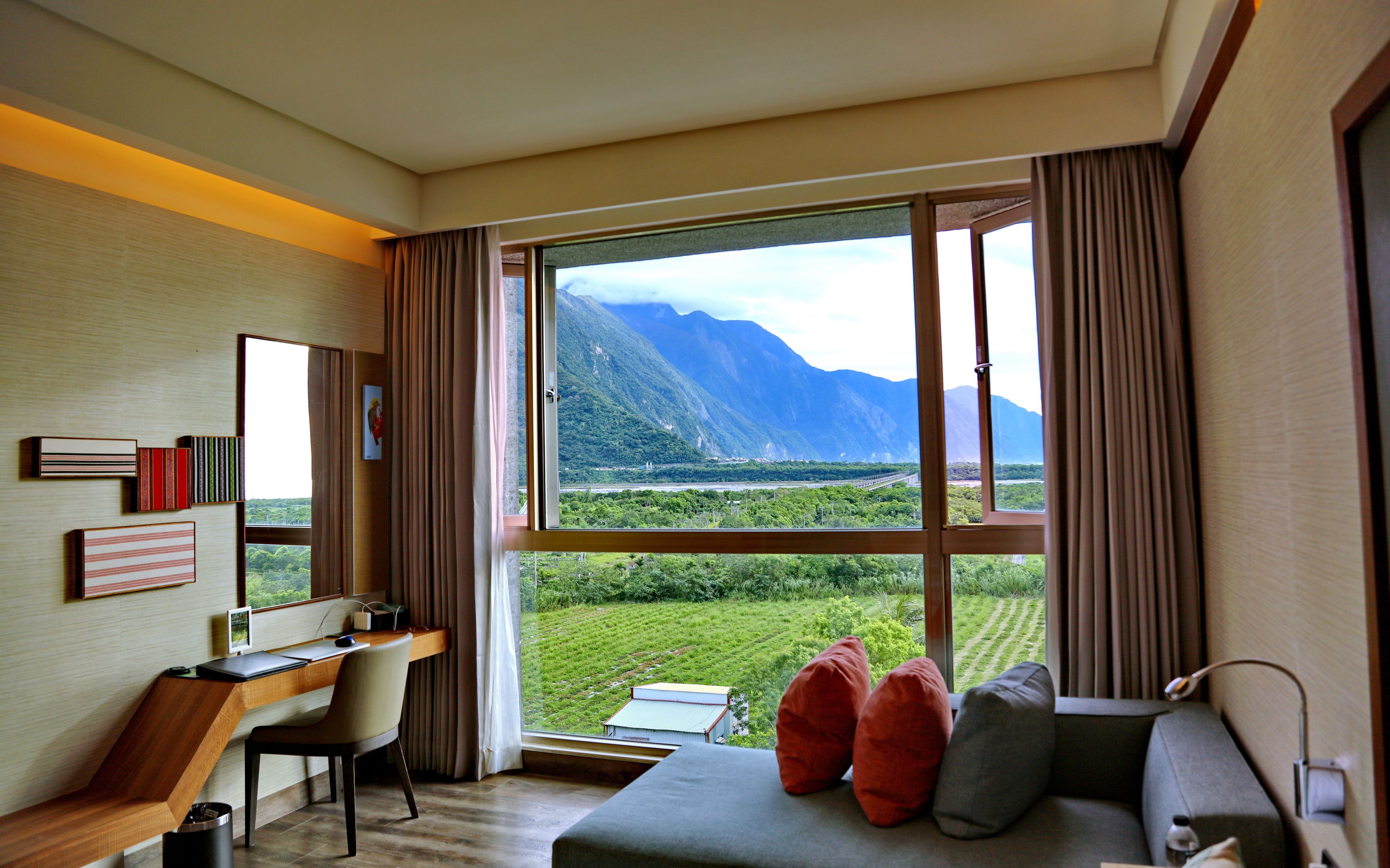 Quarto de hotel com escrivaninha, cadeira e uma grande janela com vista para uma paisagem montanhosa.