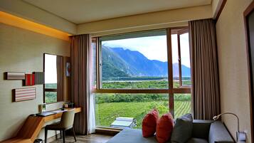 Quarto de hotel com escrivaninha, cadeira e uma grande janela com vista para uma paisagem montanhosa.