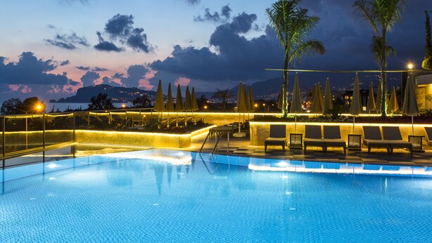 Piscina com espreguiçadeiras e guarda-sóis, cercada por uma grade e palmeiras, com um horizonte urbano ao fundo.
