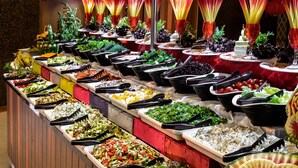 Buffet com diversos alimentos dispostos em bandejas, incluindo saladas, legumes e frutas.
