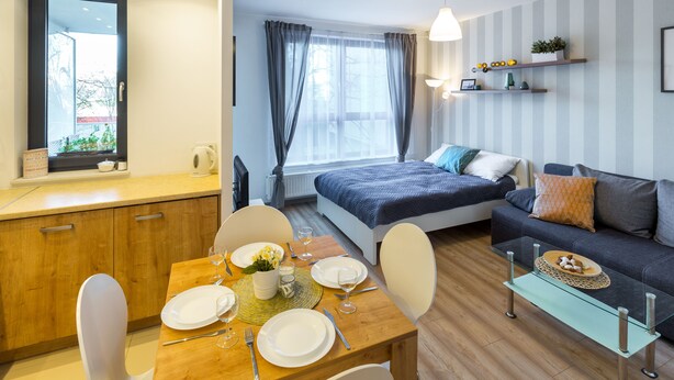 Quarto de hotel moderno com cama, sofá, área de jantar com mesa posta para dois e cozinha.
