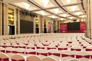Um amplo auditório com fileiras de cadeiras brancas, um palco com uma tela grande e um banner vermelho.