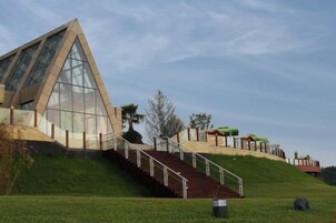 Um edifício moderno com fachada de vidro triangular, uma escadaria que leva até ele e uma área gramada em primeiro plano.