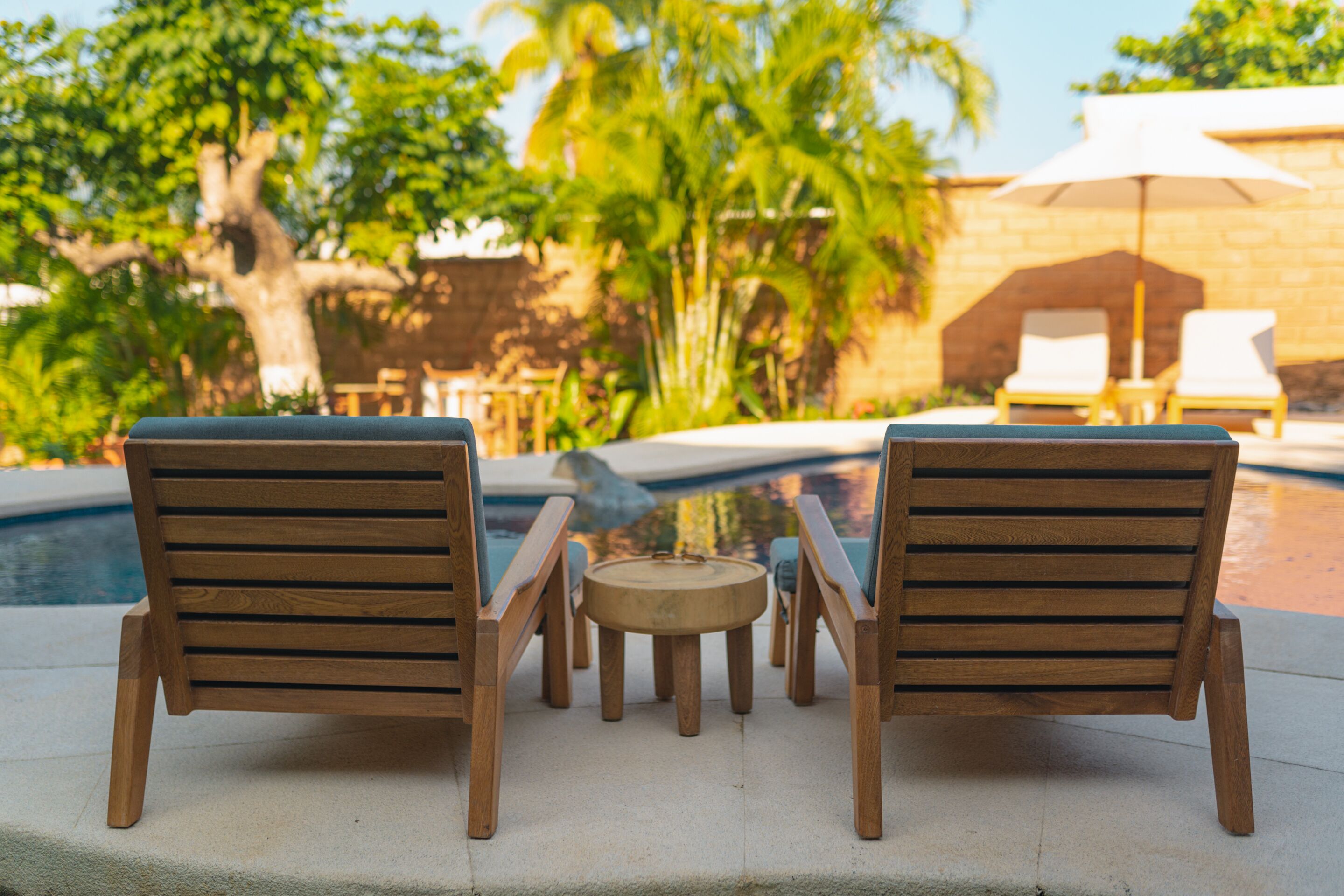 Duas poltronas de madeira com almofadas, uma mesa redonda pequena e um guarda-sol branco em um ambiente de piscina, com palmeiras e um muro de tijolos ao fundo.