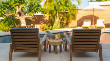 Duas poltronas de madeira com almofadas, uma mesa redonda pequena e um guarda-sol branco em um ambiente de piscina, com palmeiras e um muro de tijolos ao fundo.