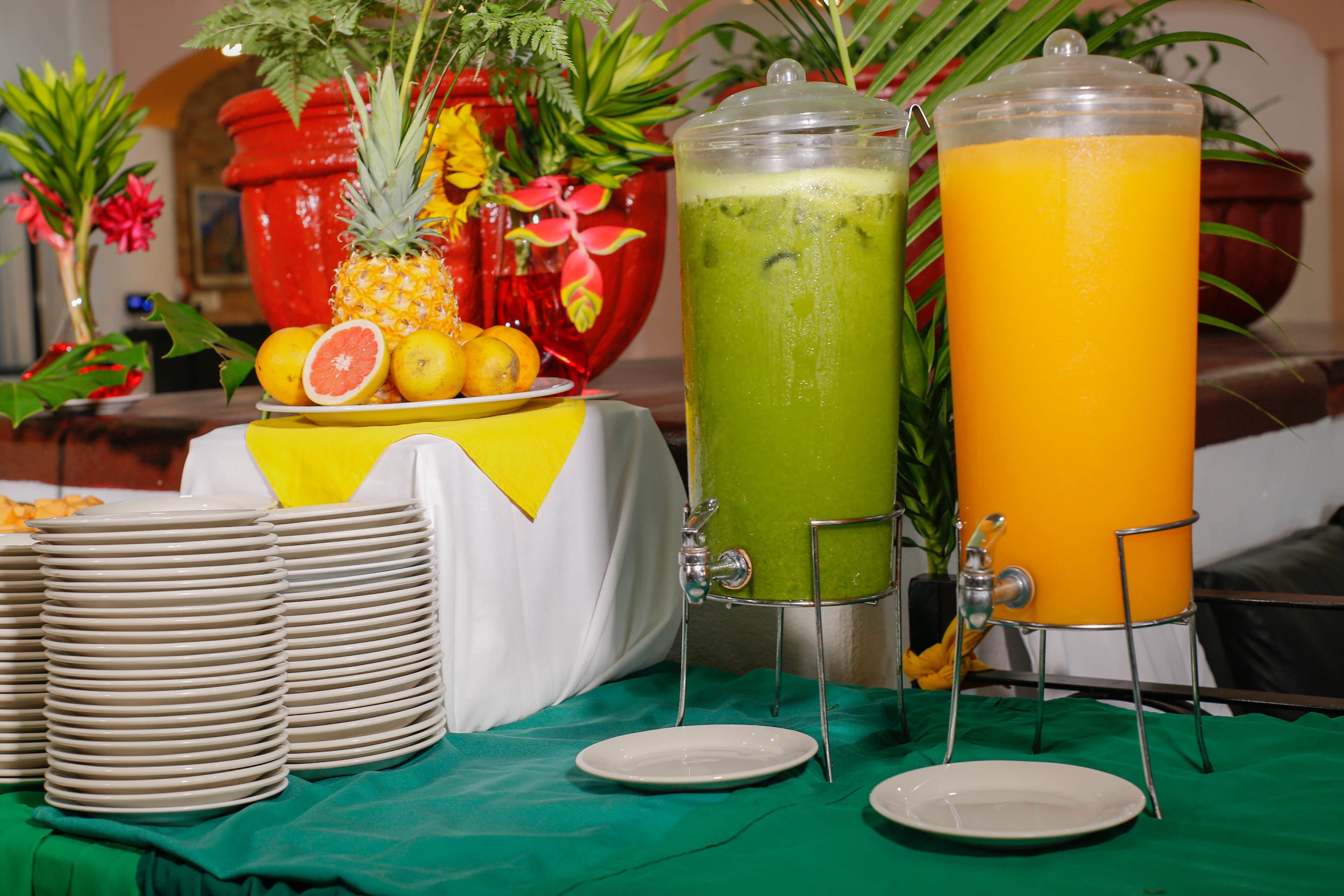 Disposição de buffet com apresentação de frutas, um dispensador de suco verde e um dispensador de suco de laranja.