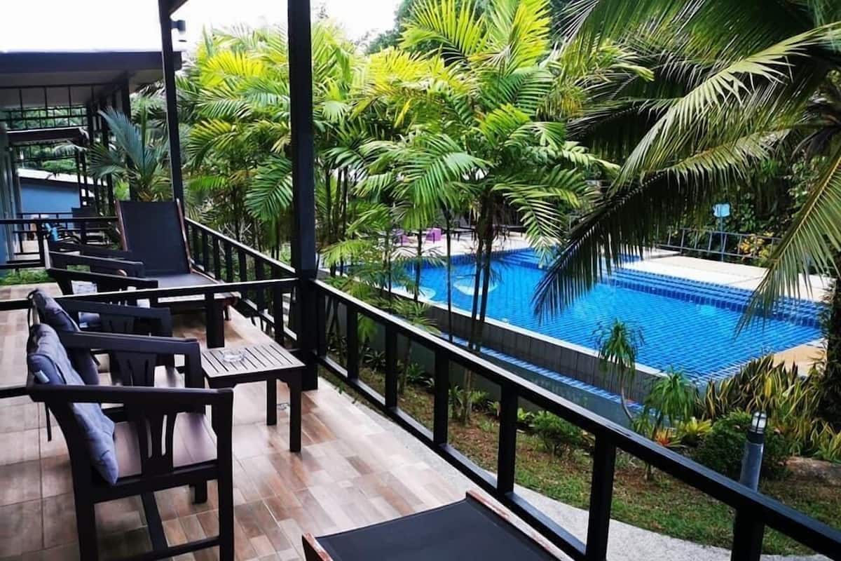 Uma varanda com piso de madeira, cadeiras com moldura preta e vista para uma piscina cercada por palmeiras.