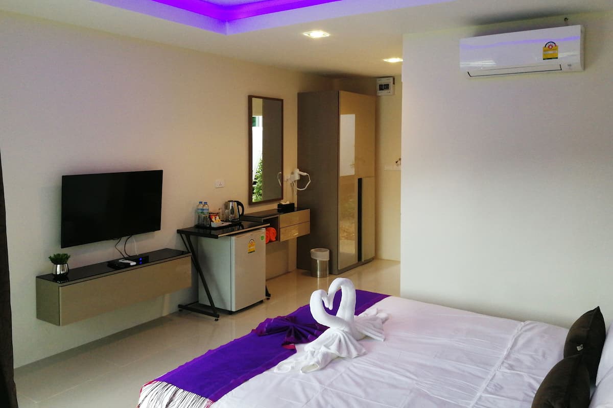 Quarto de hotel moderno com televisão de tela plana, minibar, espelho, cama com cobre-leito roxo e ar-condicionado fixado na parede.