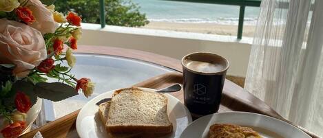 Bandeja de café da manhã com torradas, café, omelete e diversos acompanhamentos em uma varanda com vista para o mar.