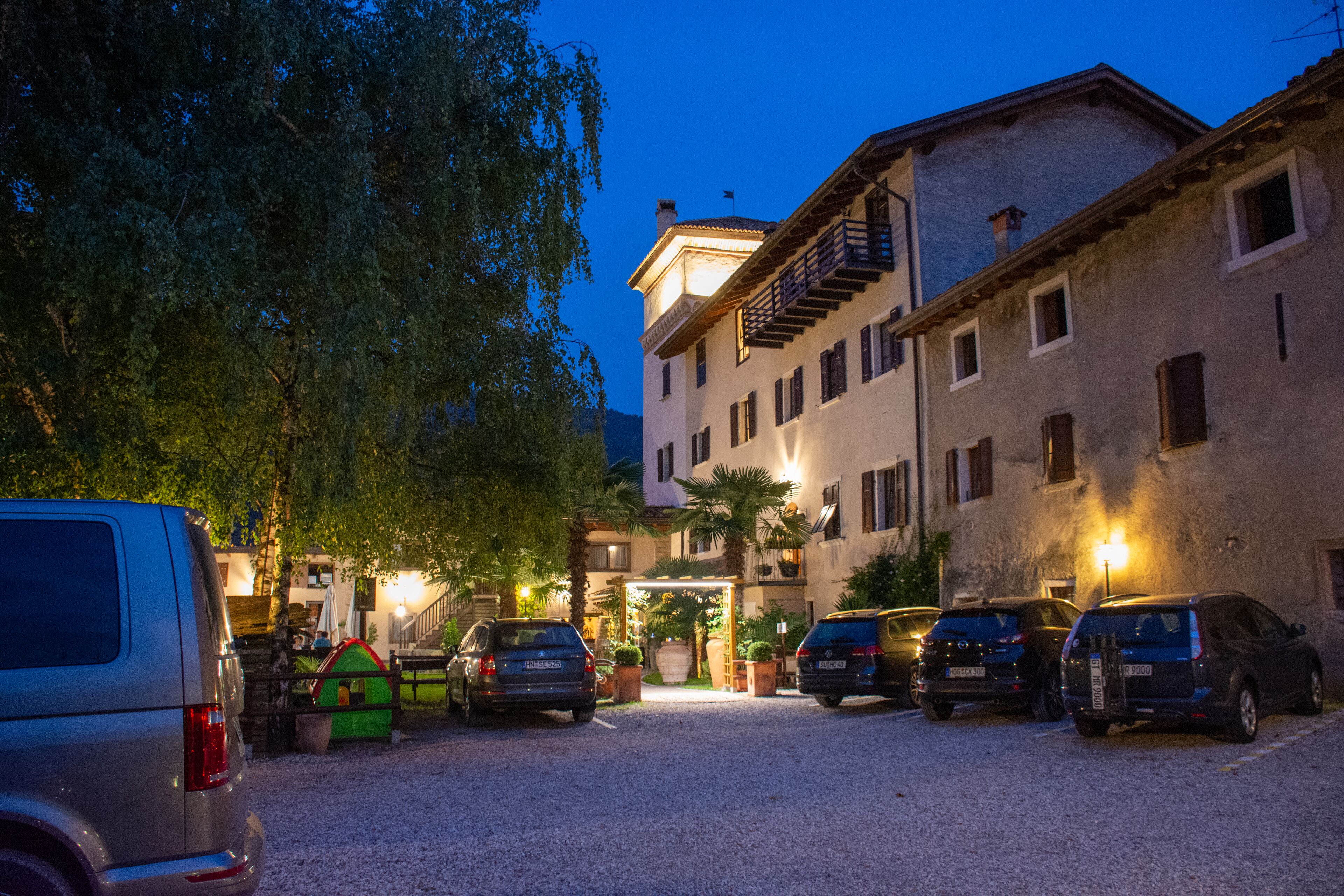 Um estacionamento com vários carros, um edifício com diversas janelas e sacadas, e uma entrada bem iluminada.