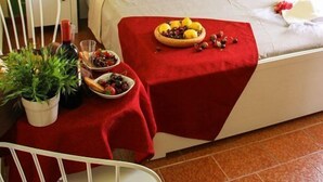 Quarto bem organizado com cama, mesas de cabeceira, uma mesa vermelha com frutas e um vaso de planta.