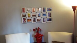 Quarto com duas poltronas brancas com almofadas estampadas, uma mesa redonda pequena e um vaso vermelho com flores. Há quadros nas paredes e um abajur.