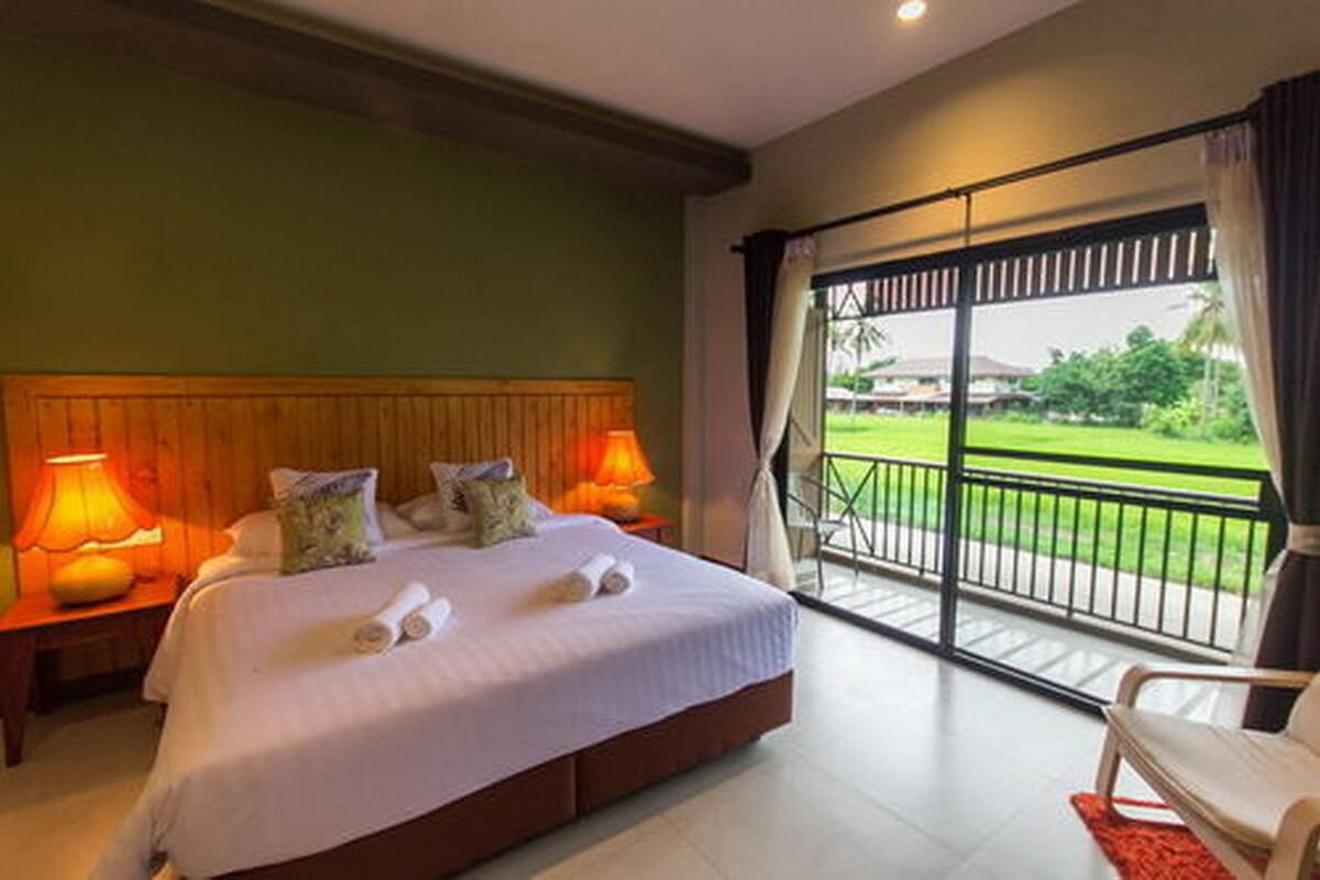 Quarto de hotel com uma cama grande, duas luminárias de cabeceira, vista para uma paisagem verde através de uma porta de correr e uma varanda com uma cadeira.