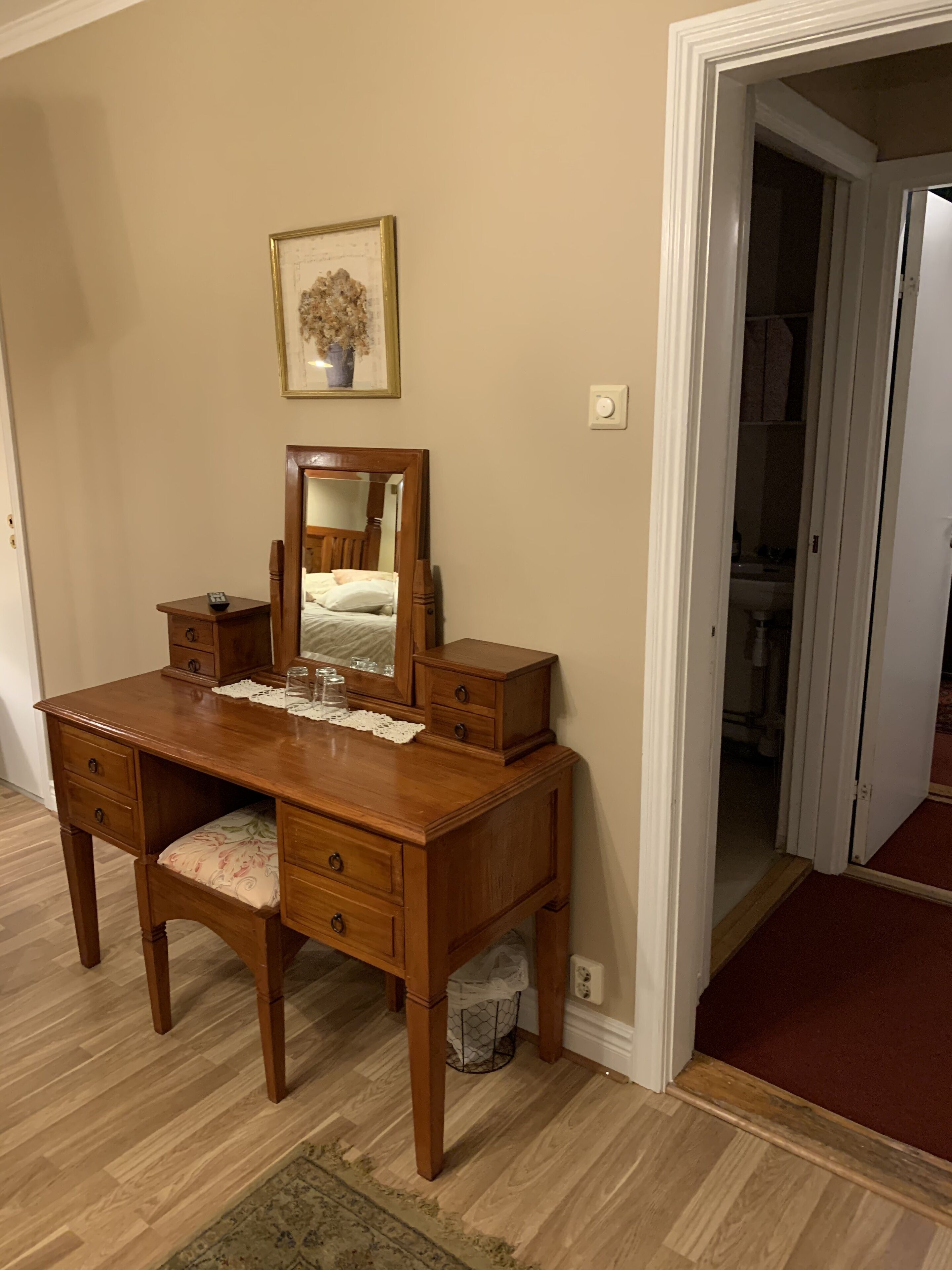 Uma cômoda de madeira com gavetas e um espelho, uma pequena mesa com um vaso e copos, e um tapete estampado sobre um piso de madeira.