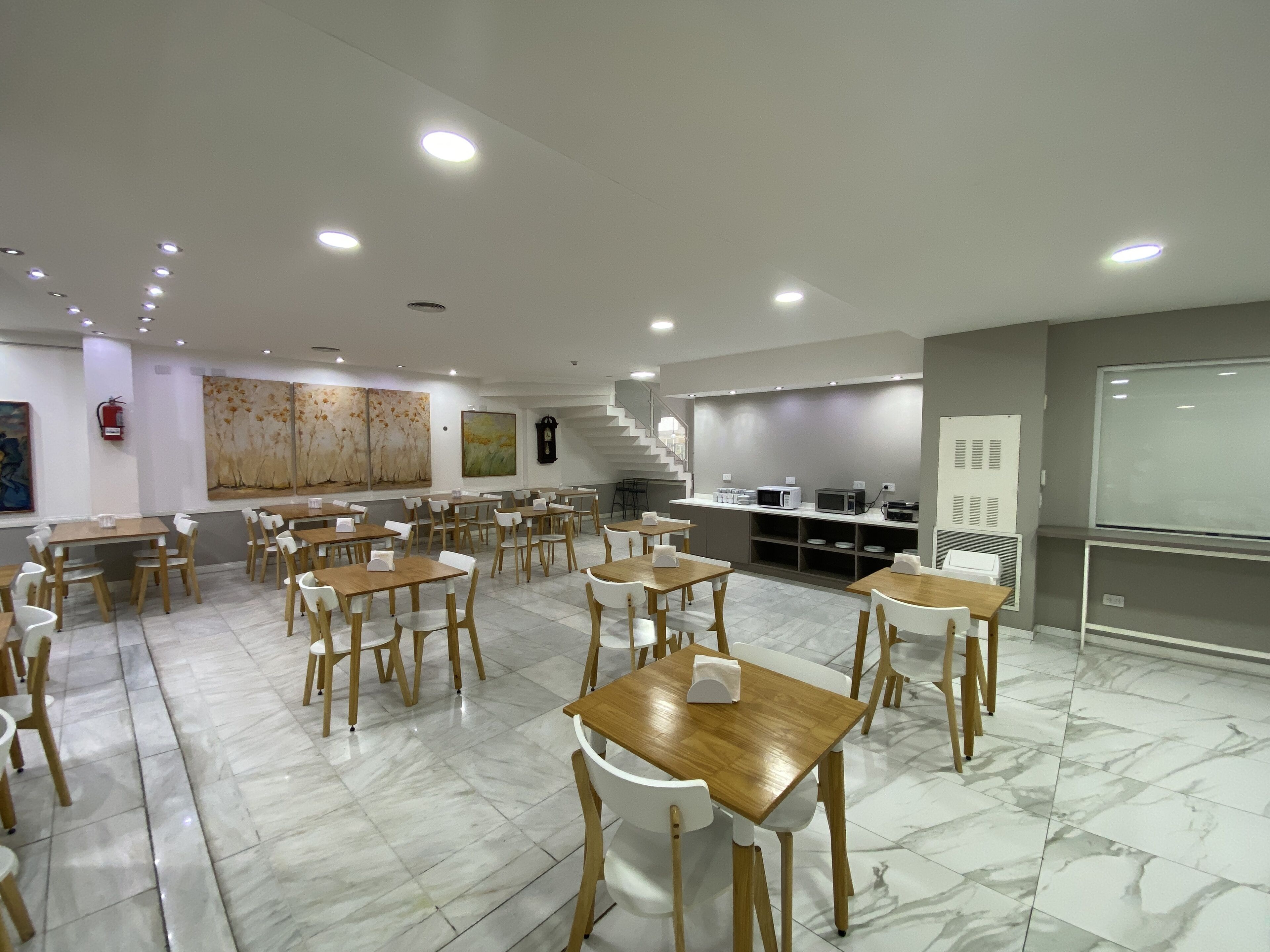Uma sala de jantar ampla com várias mesas e cadeiras, área de cozinha com eletrodomésticos e um quadro branco na parede.