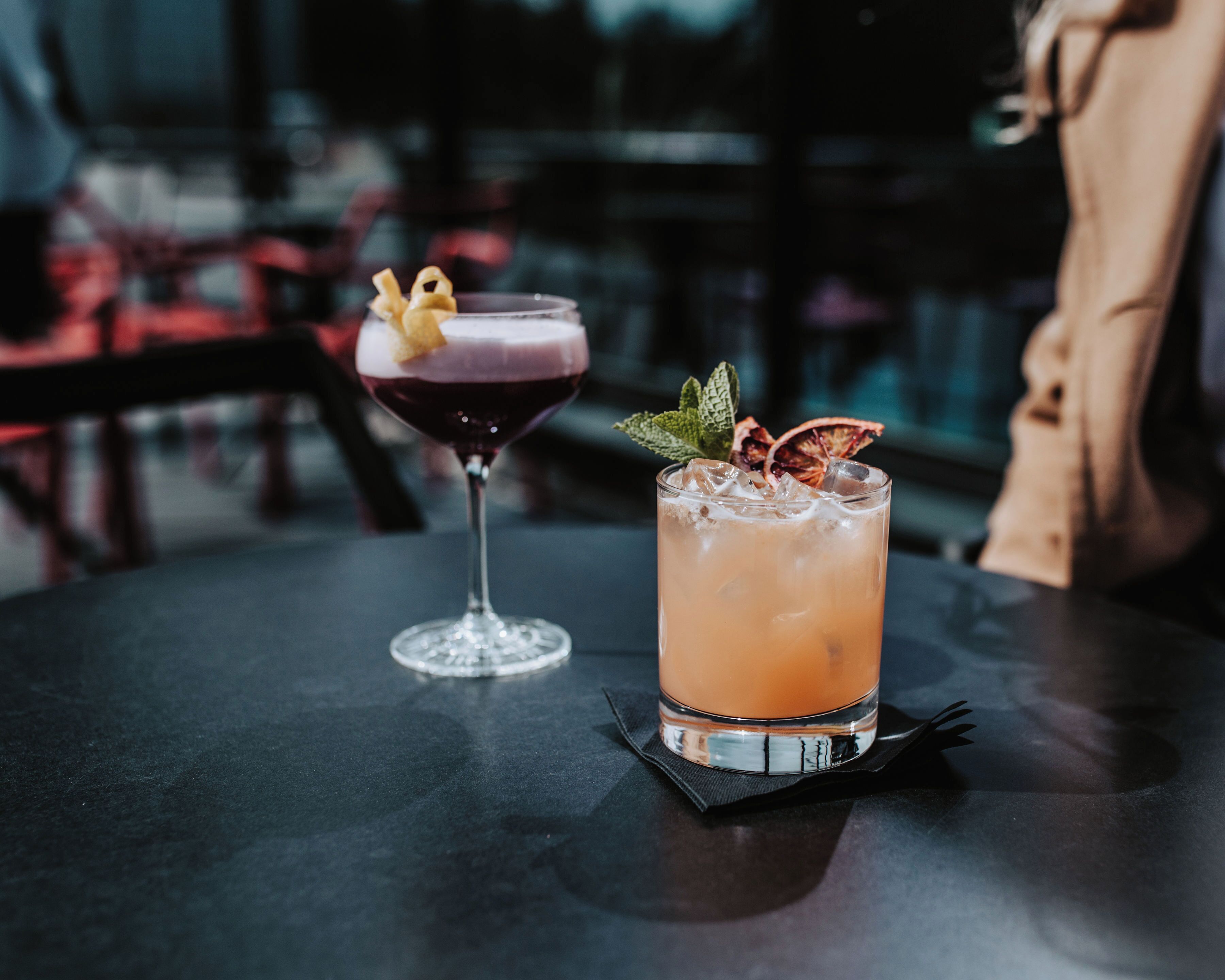 Duas bebidas sobre uma mesa com superfície escura, uma em taça martini e outra em copo highball, decoradas com folha de hortelã e uma fatia de fruta.