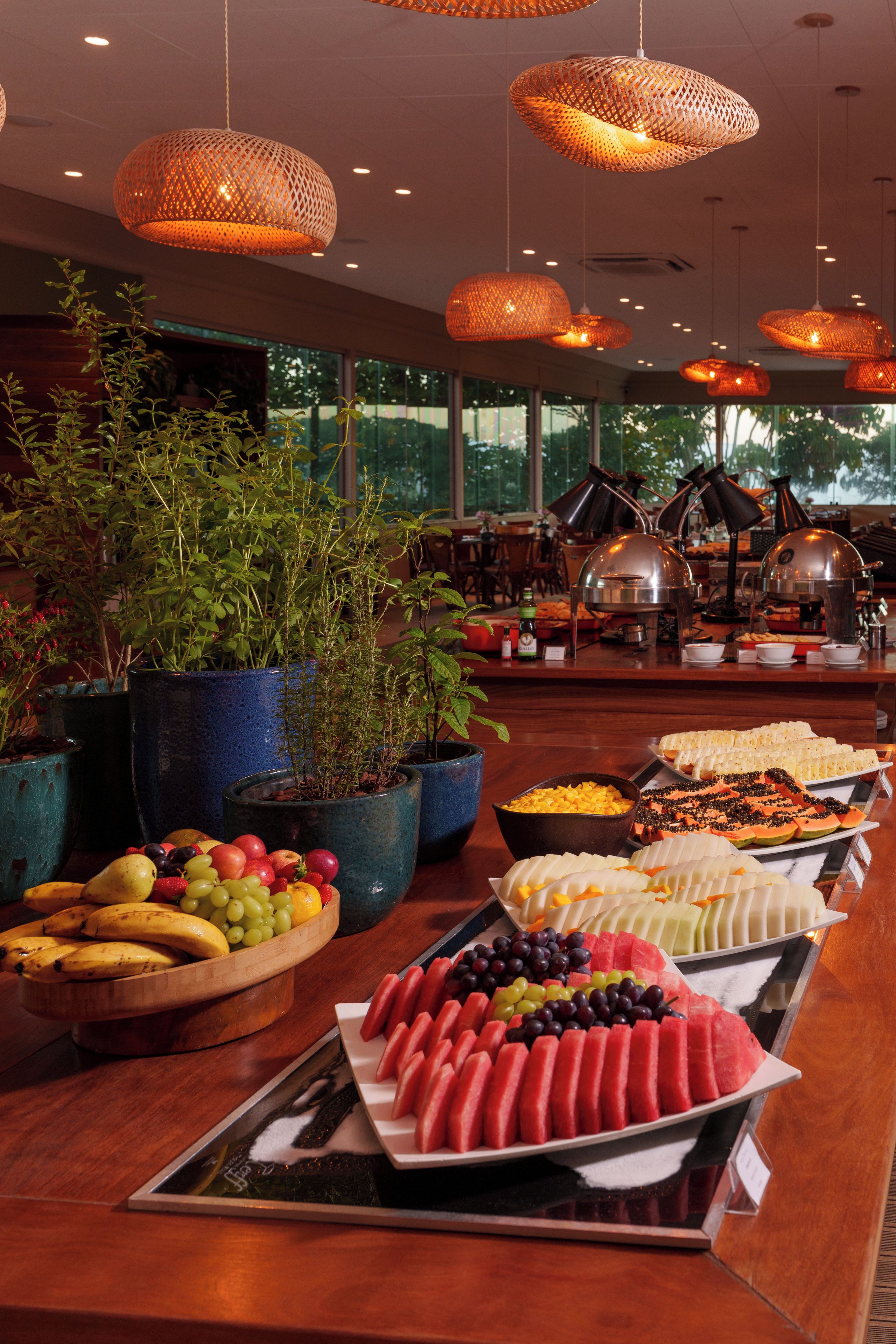 Buffet com frutas e legumes variados, plantas em vasos e um canto de café.